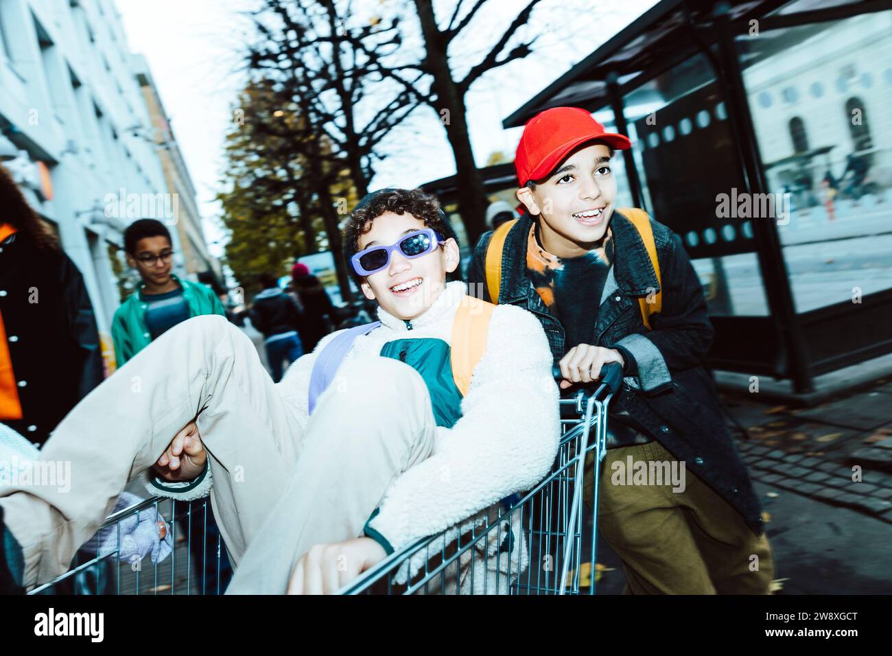 Smiling boy pushing male friend sitting in shopping cart on footpath ...