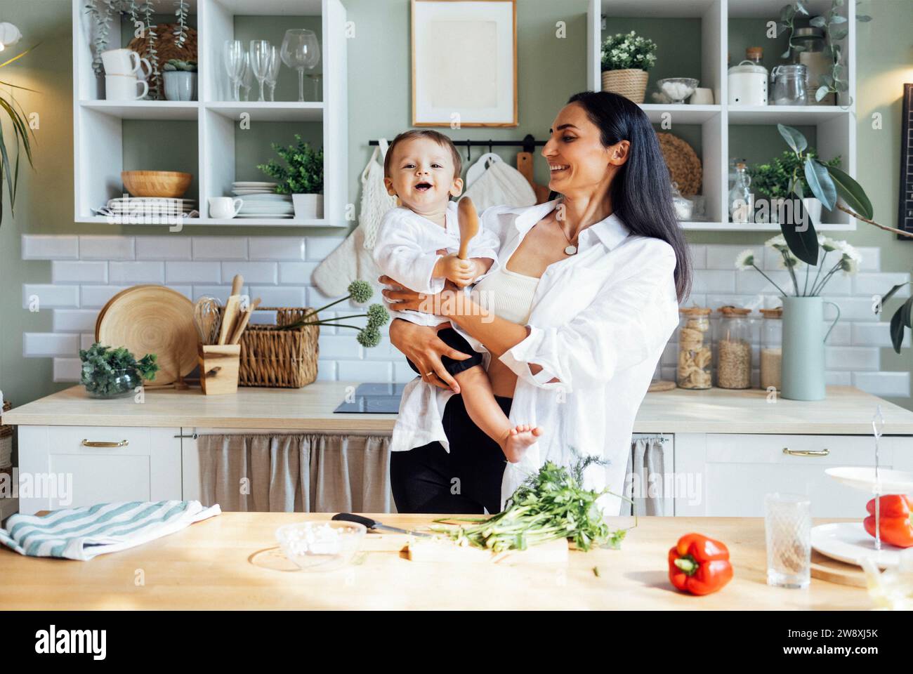 A young laughing mom holds her sweet baby in her arms in a modern kitchen. A charming vegetarian ...