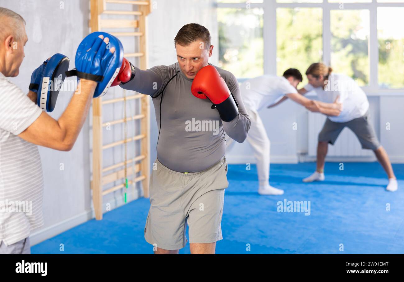 Focused man in boxing gloves practices punches with man in mitts in gym ...
