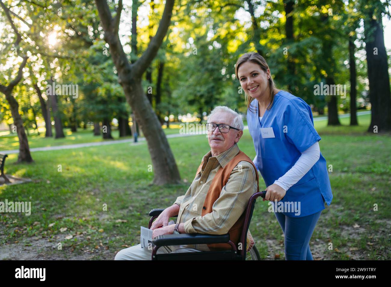 Smiling healthcare worker walking with senior man sitting in wheelchair ...