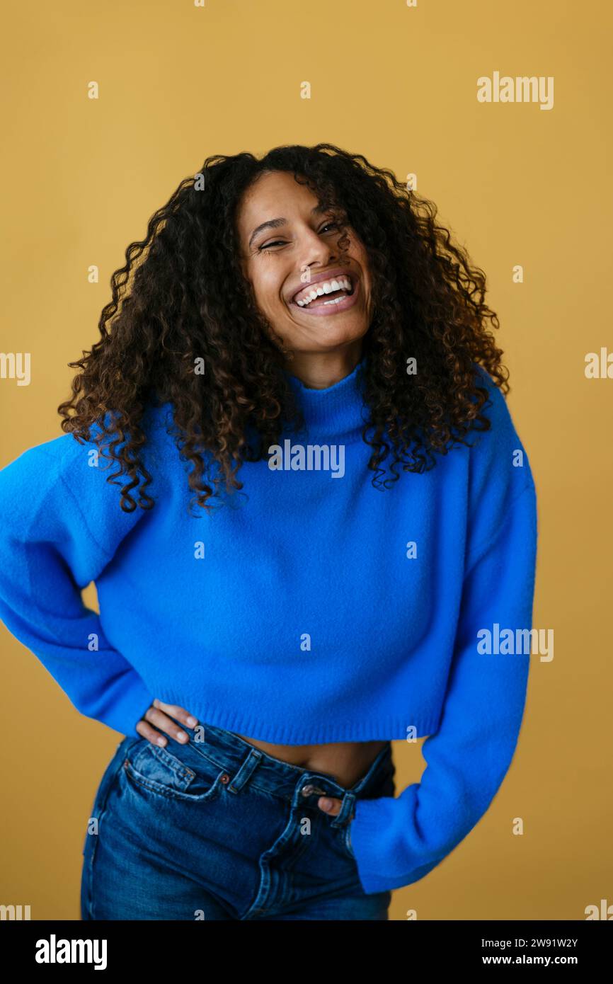 Happy woman standing with hand in pocket against yellow background ...
