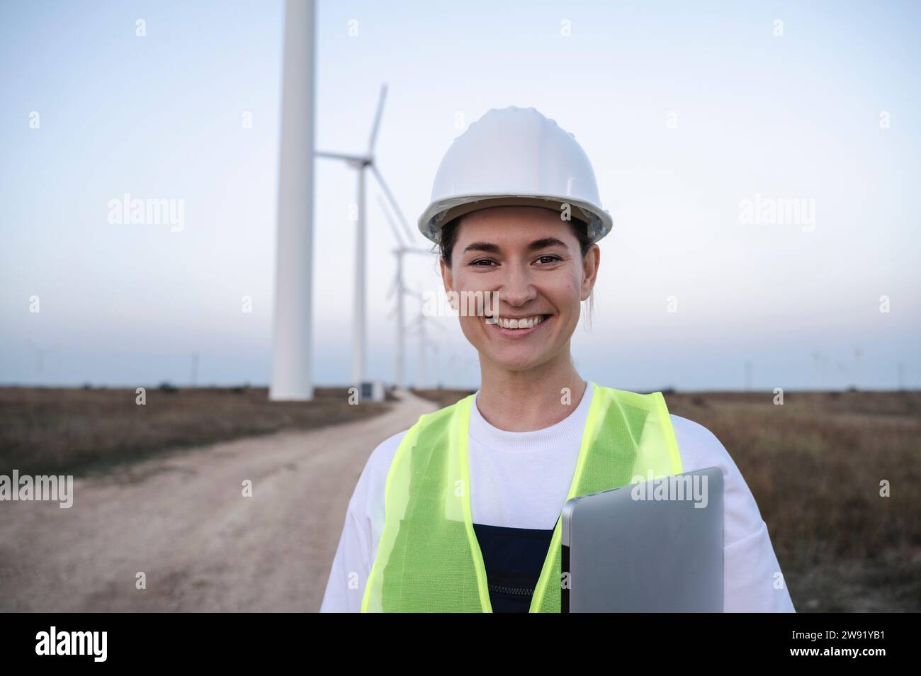 Happy engineer wearing hardhat standing with laptop in front of wind ...