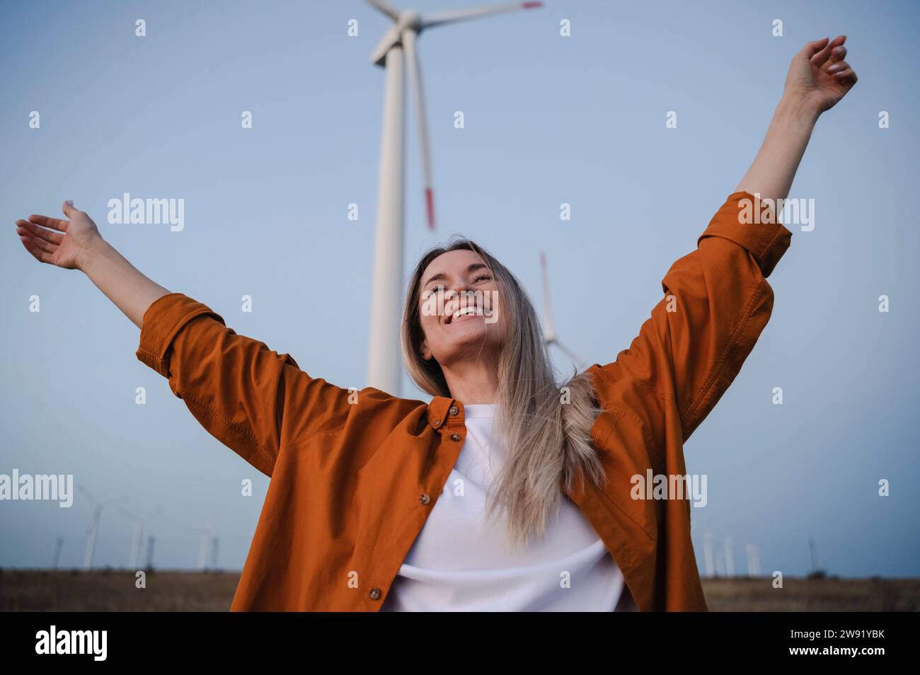 Happy woman with arms raised and eyes closed standing in front of wind ...