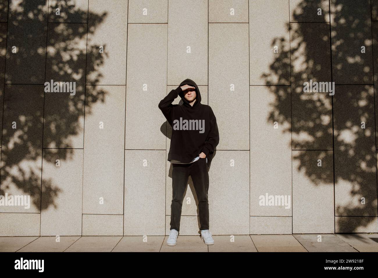 Boy with hand in pocket shielding eyes and standing in front of wall ...