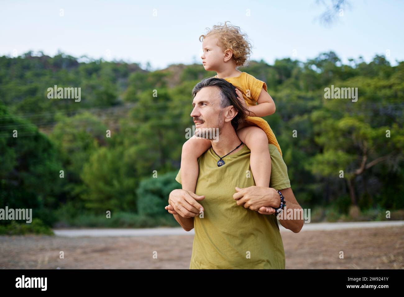 Contemplative father carrying daughter on shoulders in front of trees ...