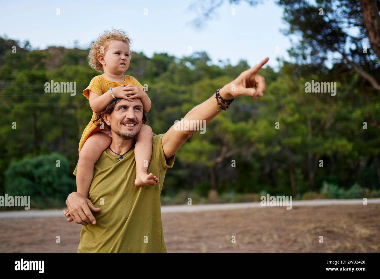 Smiling man pointing and carrying daughter on shoulders in front of ...