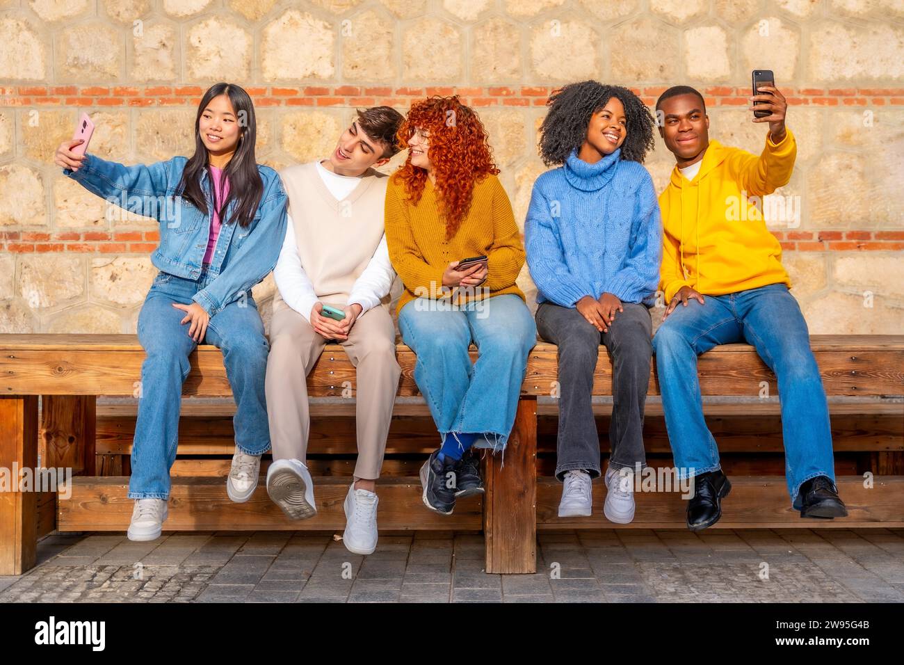 Multi-ethnic young friends taking selfies with the mobile phone sitting on a wood bench in the ...