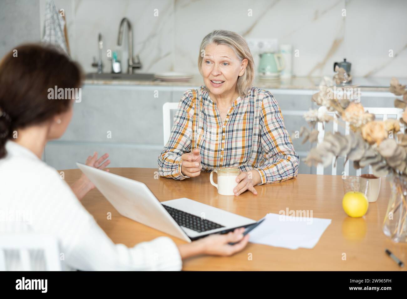 Positive old woman sitting at the kitchen-table face to face with ...