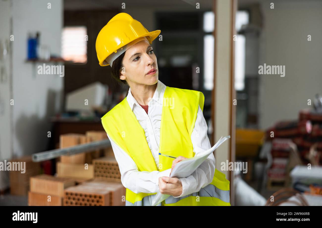 Portrait of woman builder in yellow vest and hardhat filling documentation during repair works ...