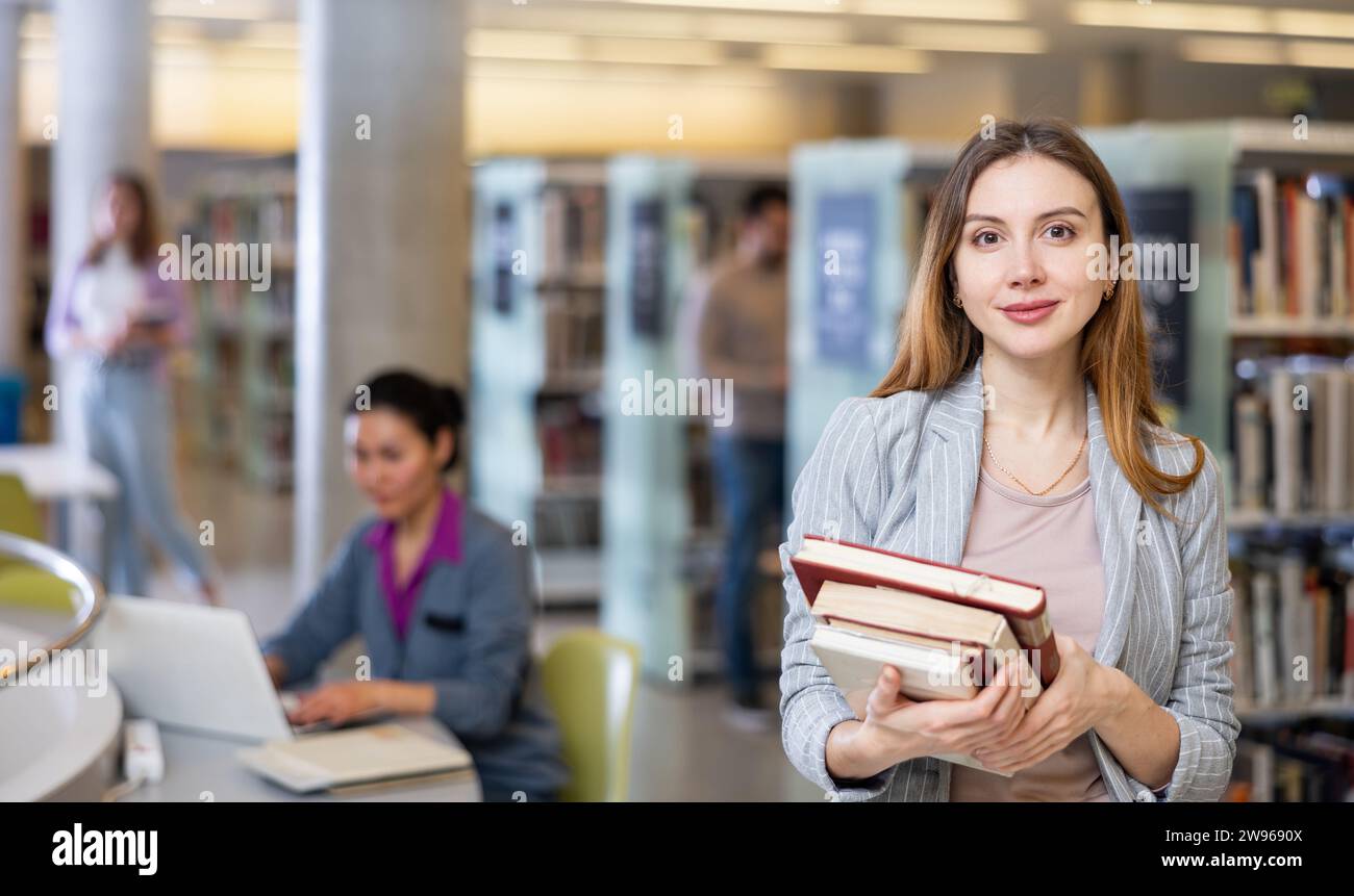Smiling young woman with stack of books standing in library Stock Photo ...