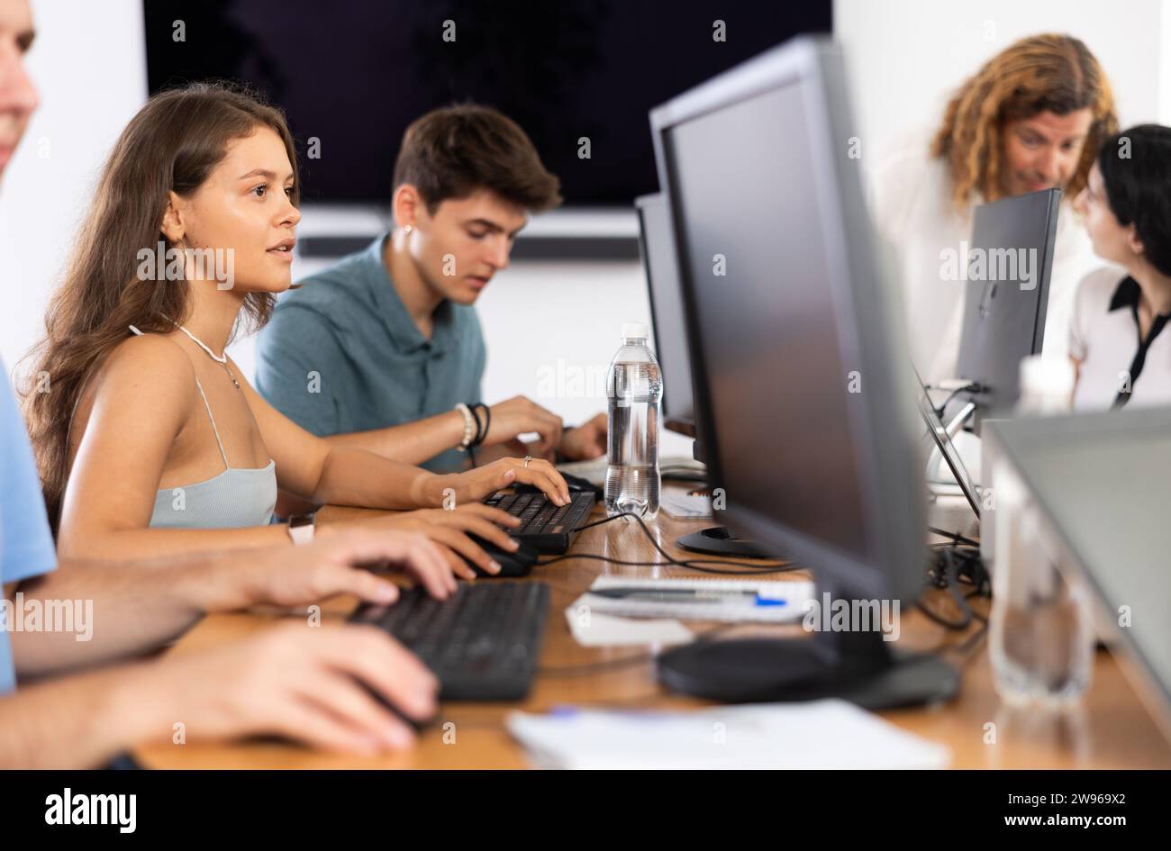 Female student of computer courses attentively looks at monitor screen, types on keyboard and ...
