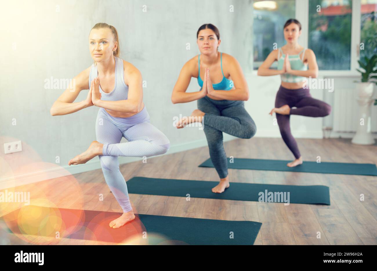 Sedulous women doing tree pose of yoga on black mat in light gym room ...