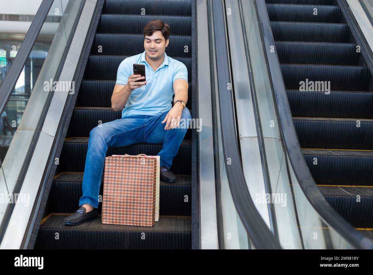 Happy young indian man sitting, riding escalator and using smart phone ...