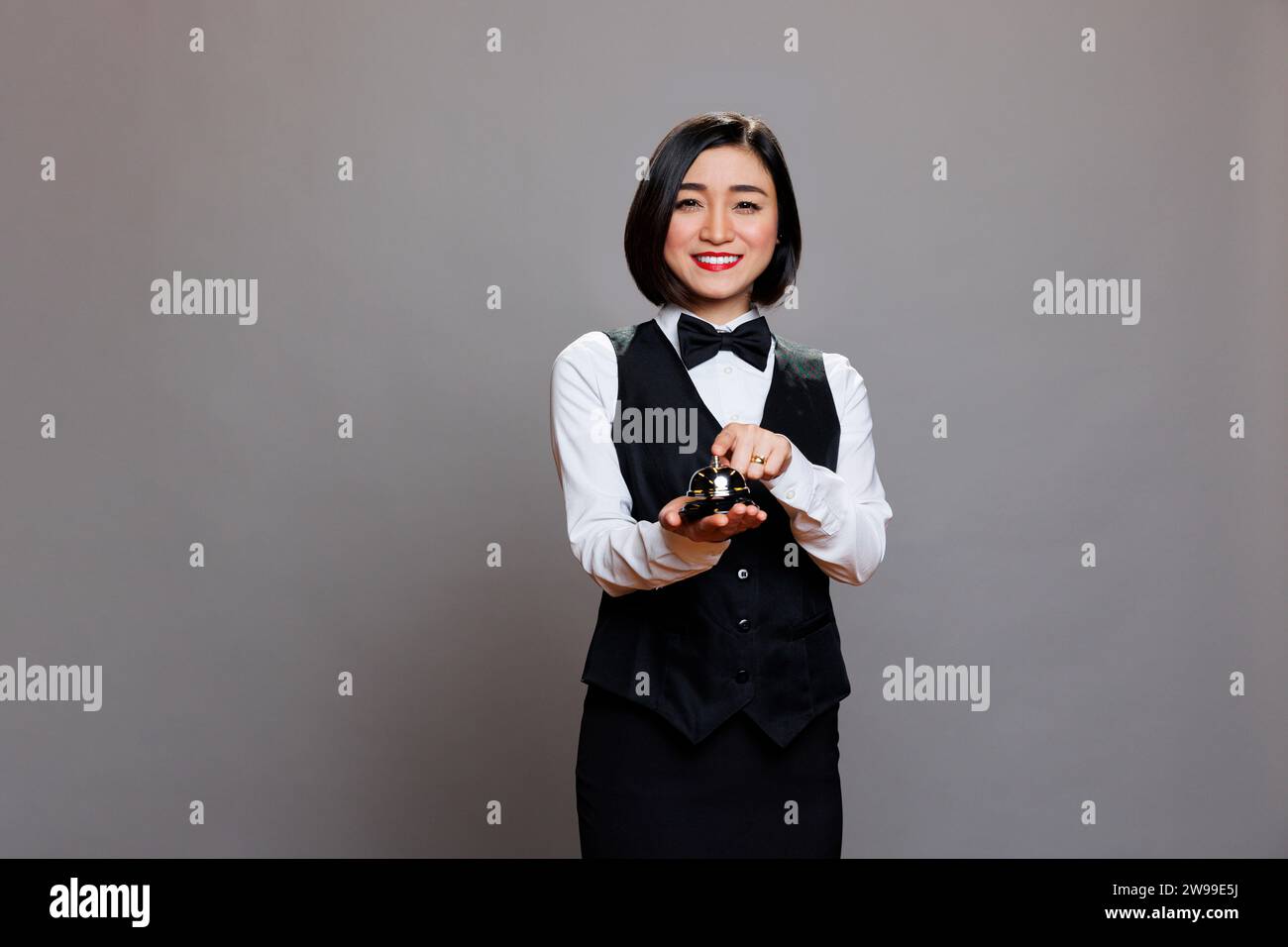 Smiling asian restaurant receptionist in uniform ringing dining bell ...