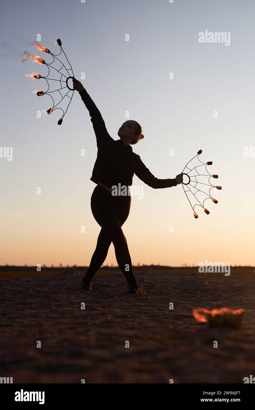 Full length portrait of female artist dancing with fire fans outdoors ...
