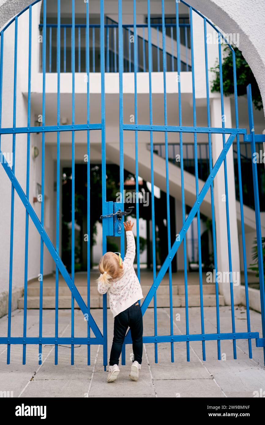 Little girl reaches for the lock on the metal arched gate of the house ...
