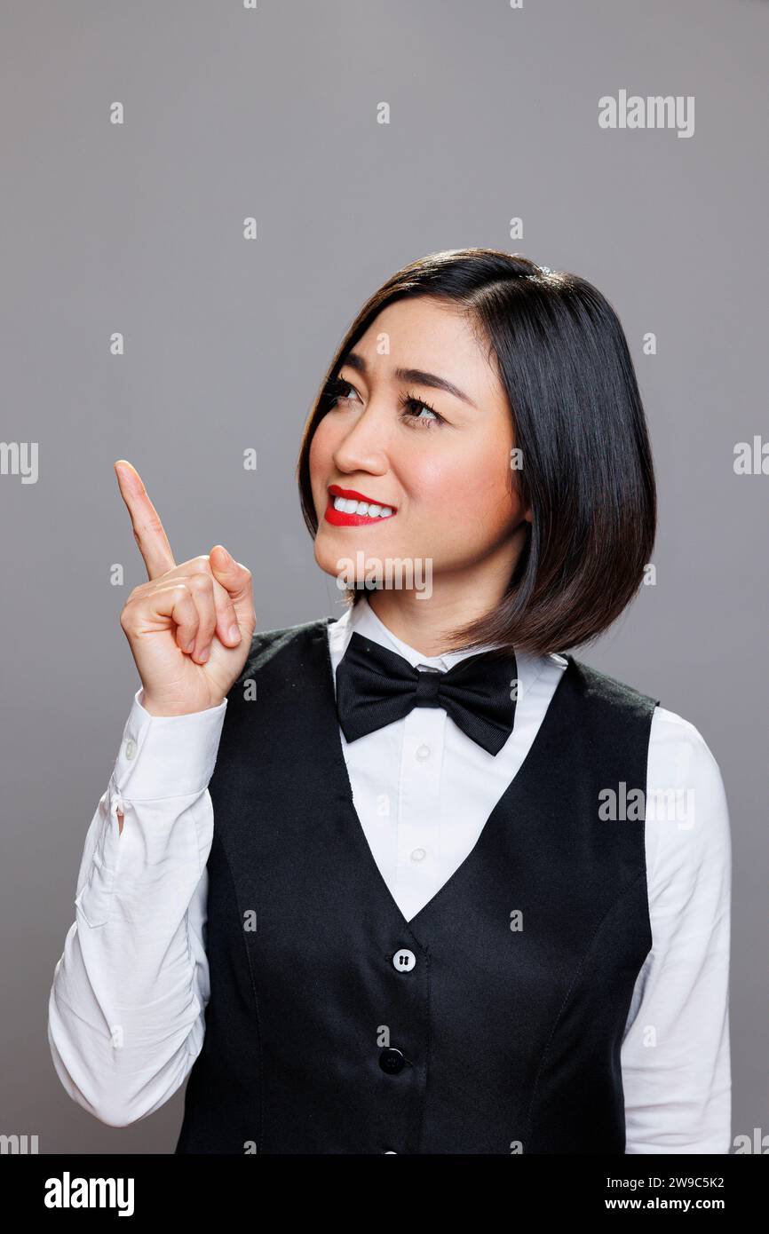Smiling asian woman receptionist showing upwards and looking to side ...