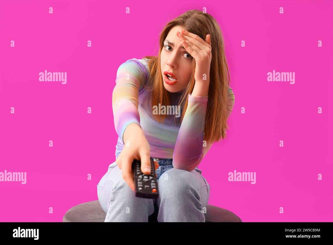 Confused young woman with remote control watching TV on pink background ...