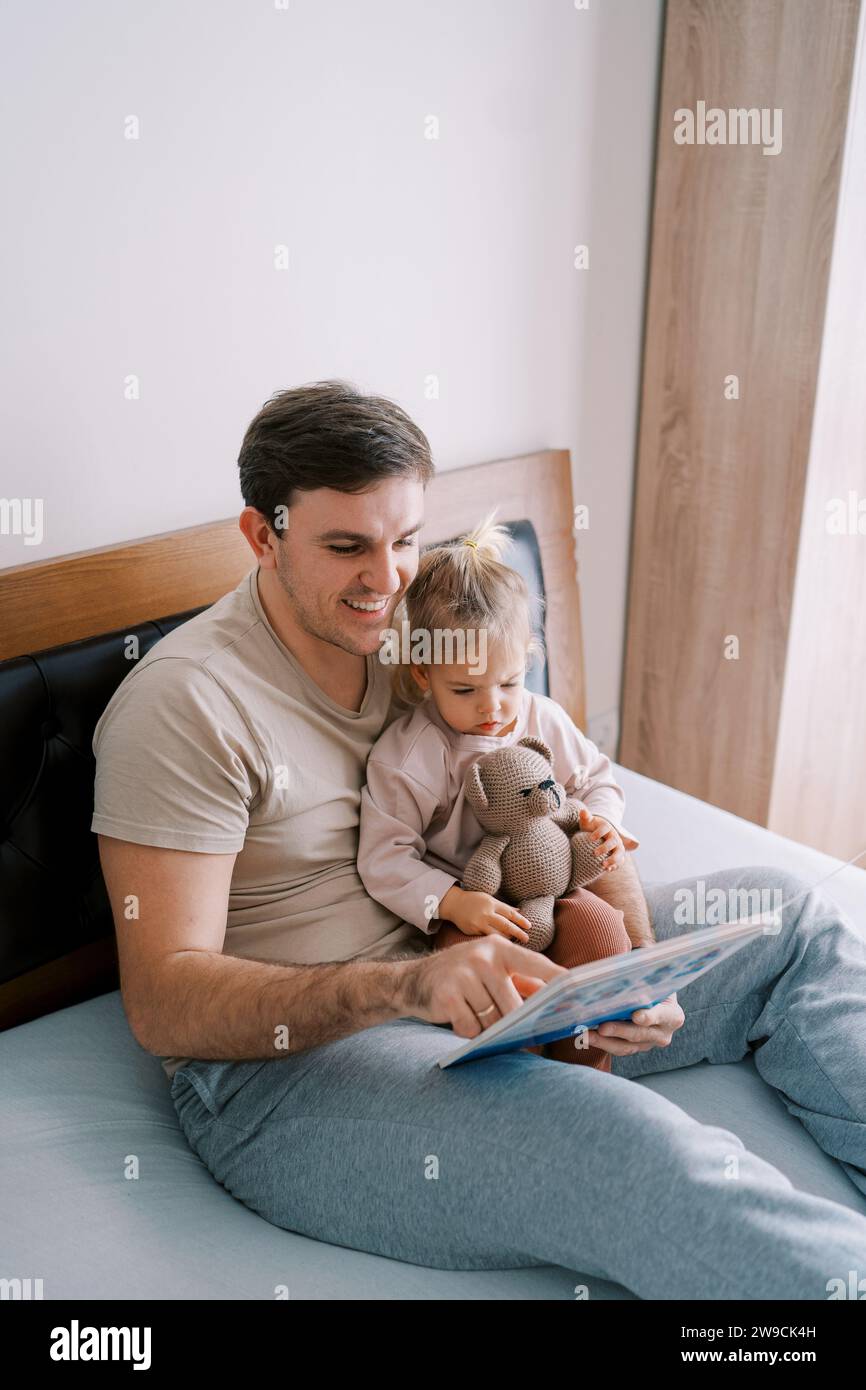 Smiling dad reading a book to a little girl sitting with a teddy bear on his lap on the bed ...