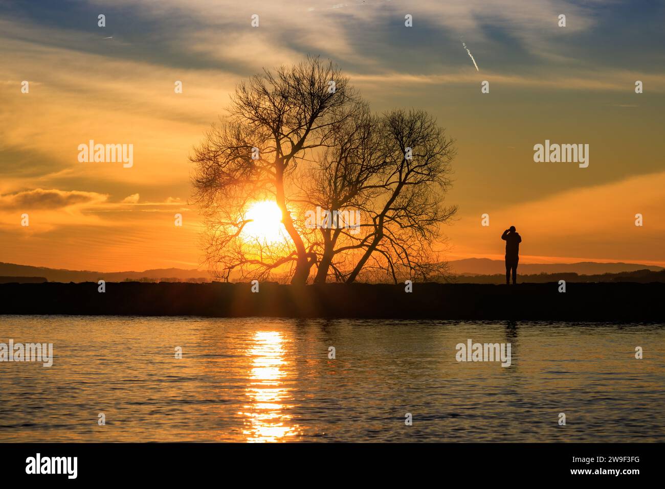 A man wearing a blue shirt taking a picture of a sun setting over the ...