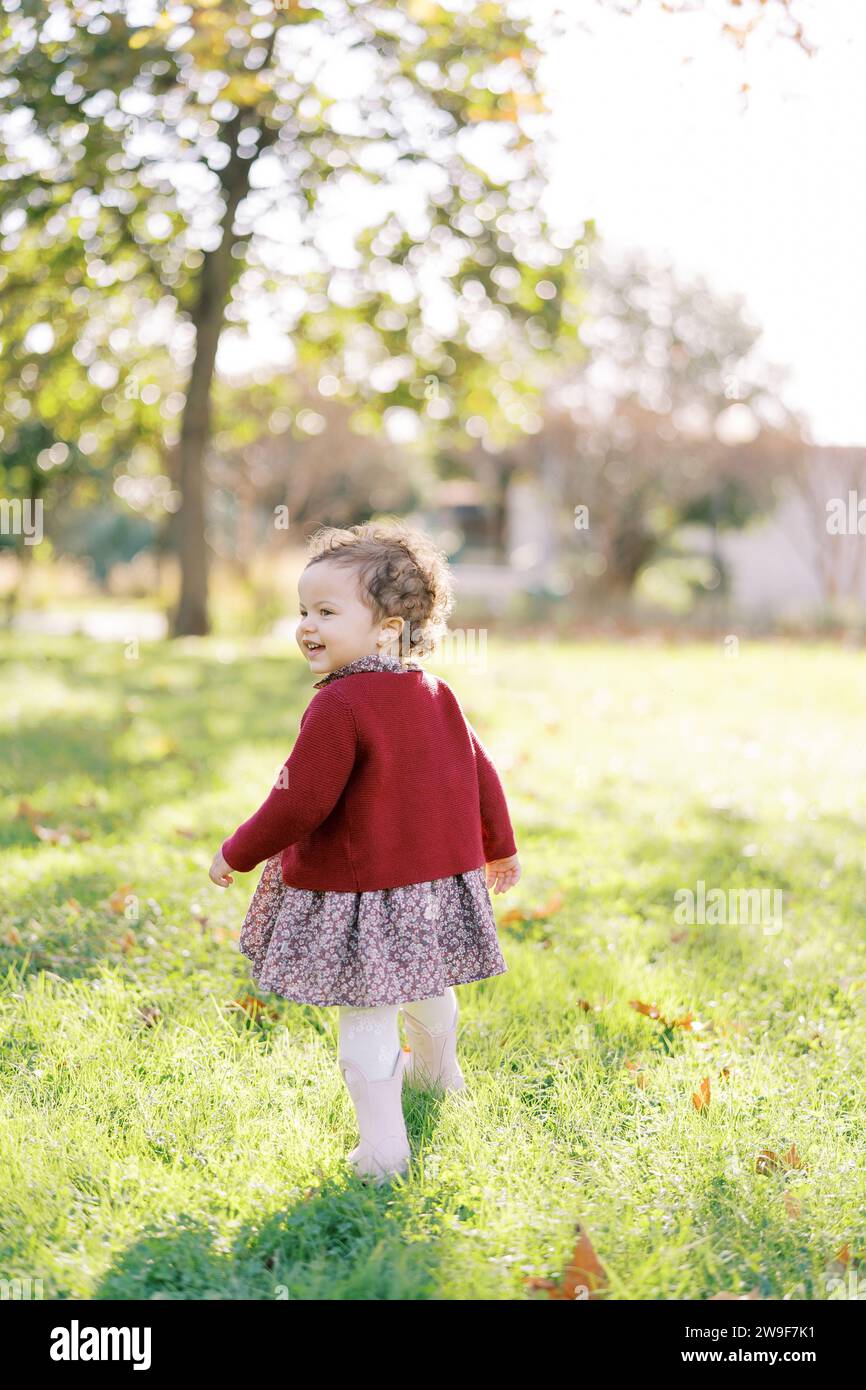 Little smiling girl walks among the fallen leaves, turning around. Back ...