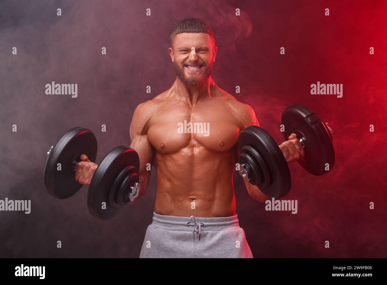 Emotional young bodybuilder exercising with dumbbells in smoke on color ...