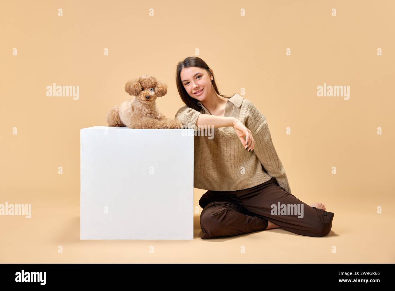 Full length portrait of young attractive lady sitting near white cube ...
