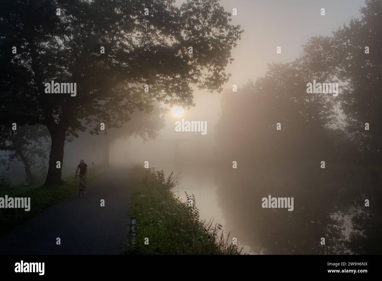 A solitary cyclist embarks on a journey along a riverside path shrouded ...