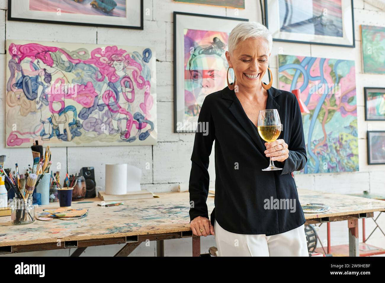 cheerful mature female artist with wine glass smiling in art studio ...