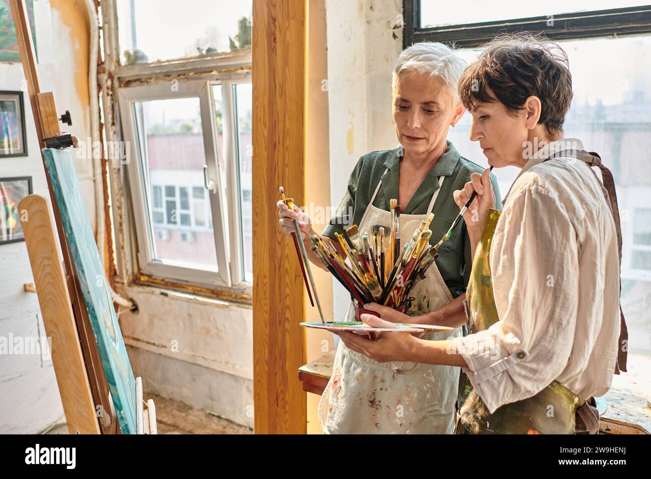 skilled female painter helping mature woman to choose paintbrush during ...