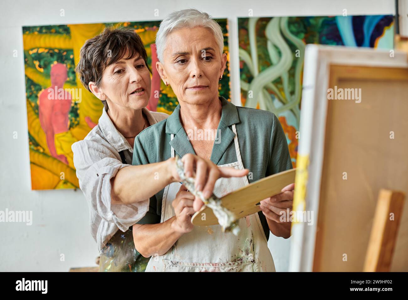 skilled female artist pointing at easel near mature woman during master ...