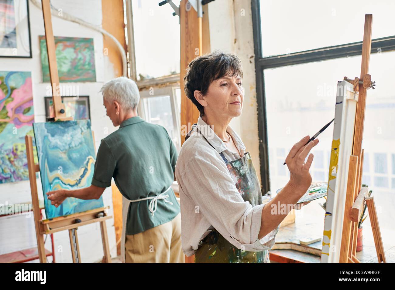 inspired brunette mature woman painting on easel near female friend in ...