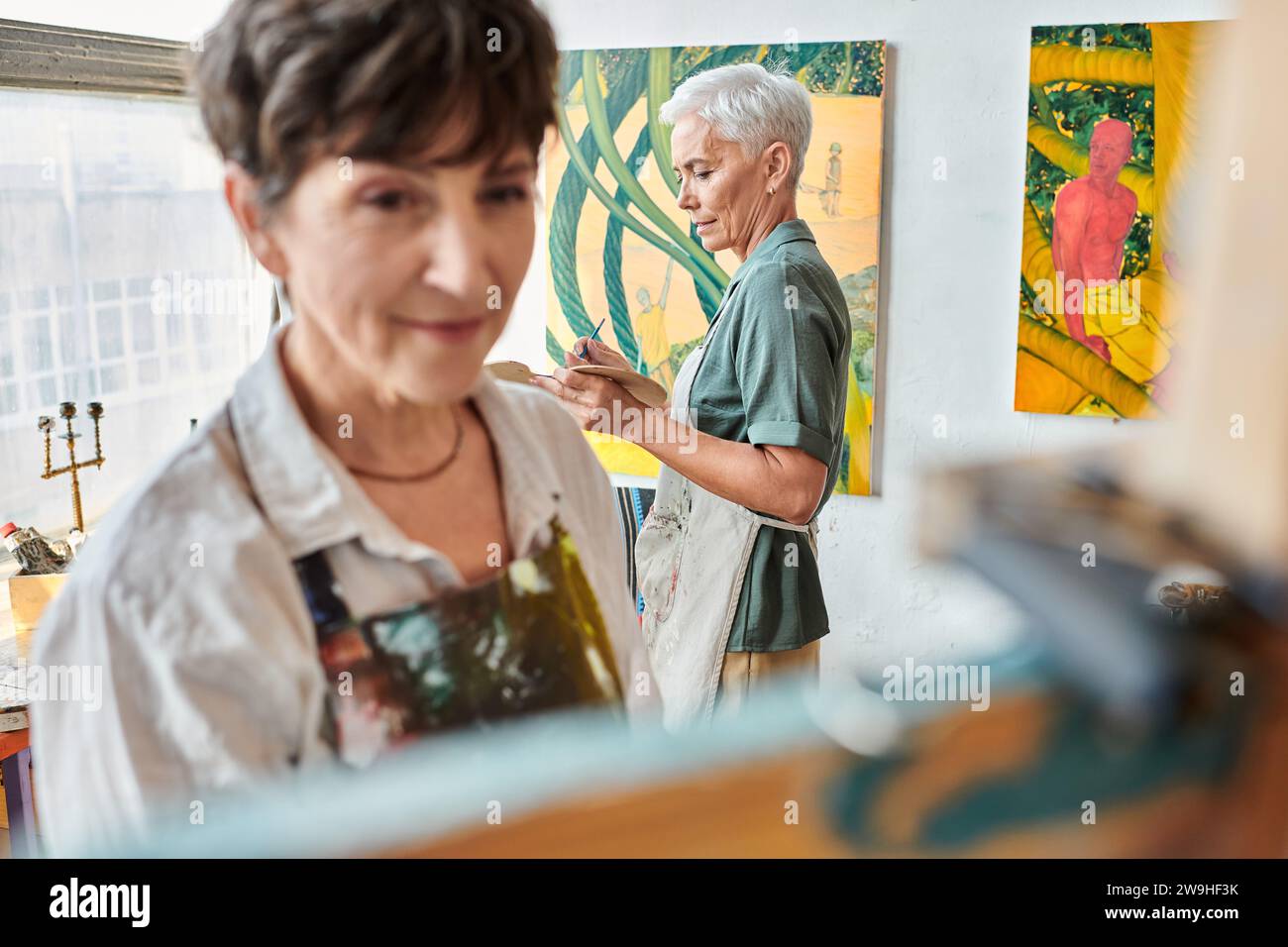 blurred female artist near easel and mature woman painting in art ...