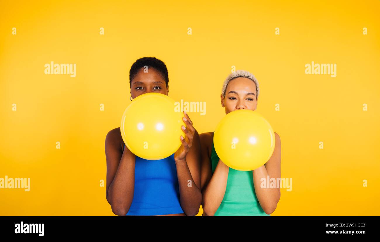 Two young women blowing a yellow balloon in front of yellow background ...
