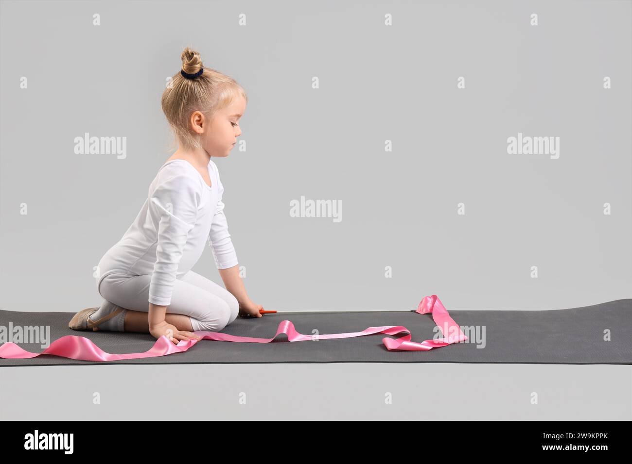 Cute little girl with ribbon doing gymnastics on mat against light ...