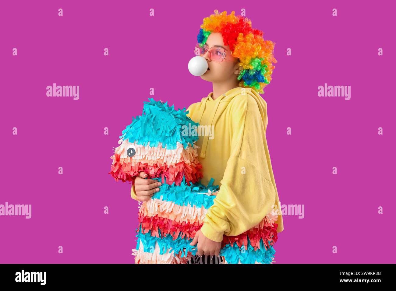 Trendy girl in clown costume with pinata blowing bubble gum on purple ...