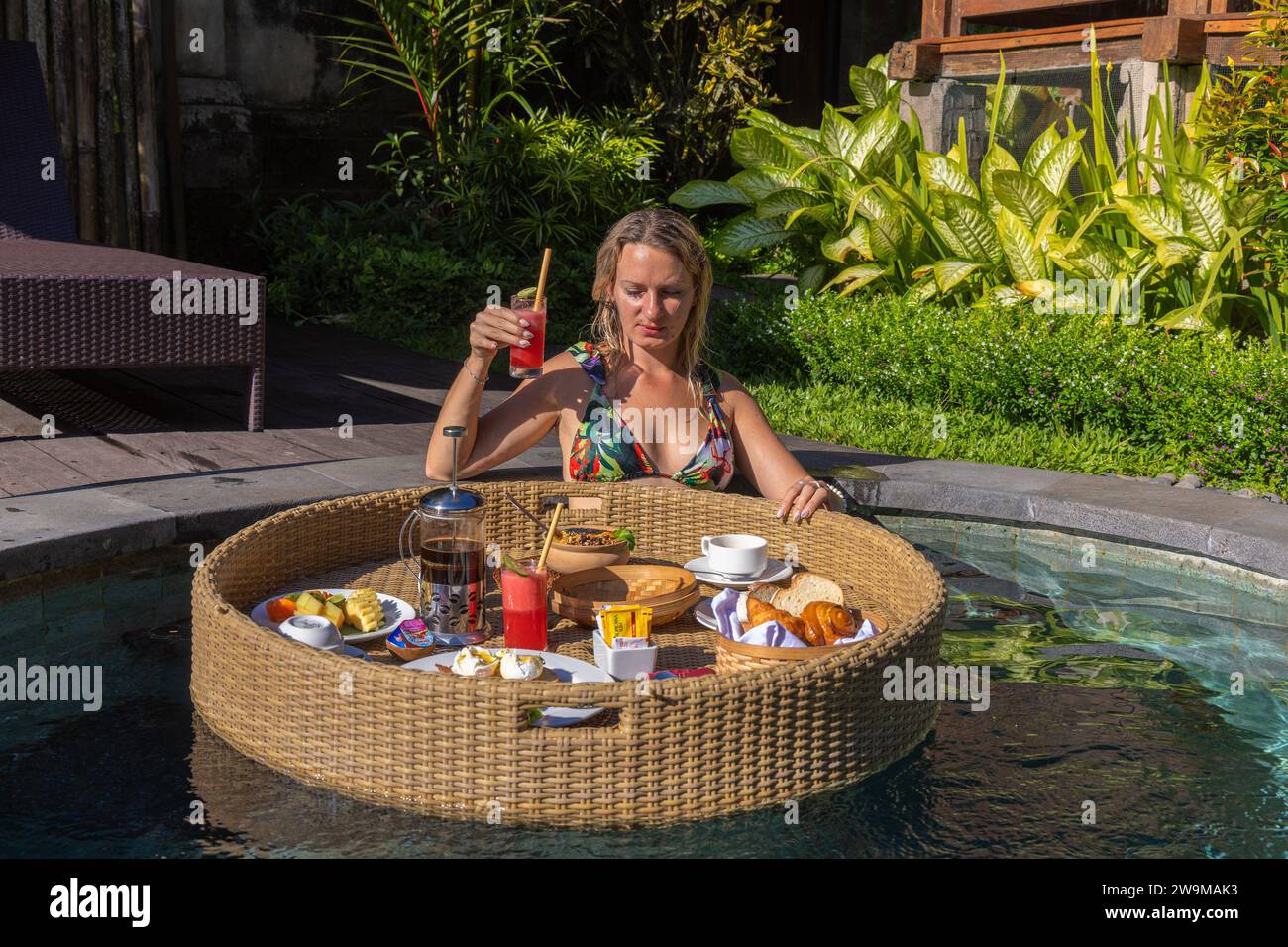 A young woman having breakfast on a floating basket in a swimming pool ...