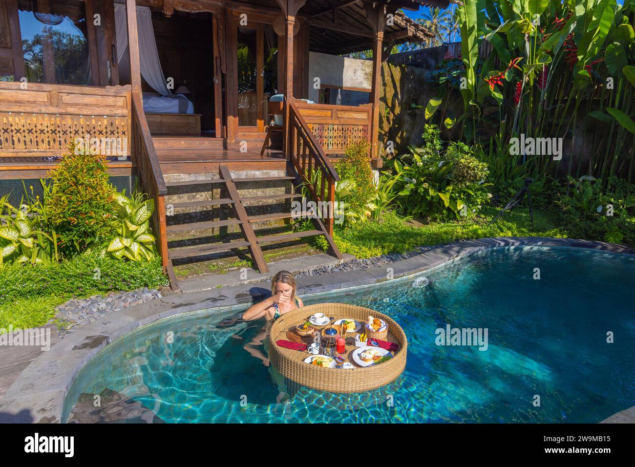 A young woman having breakfast on a floating basket in a swimming pool ...