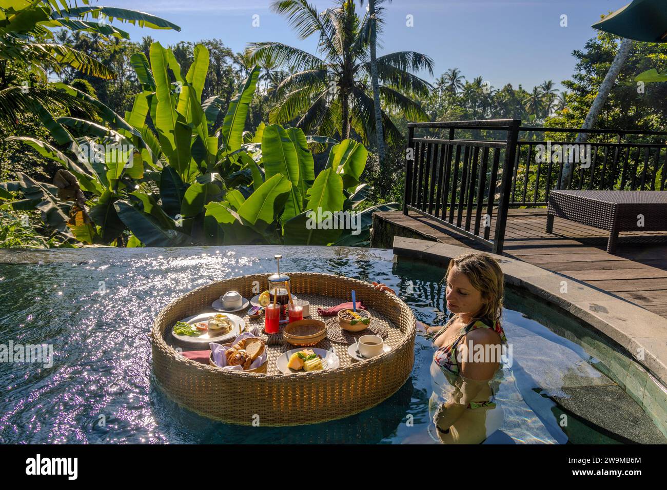 A young woman having breakfast on a floating basket in a swimming pool ...
