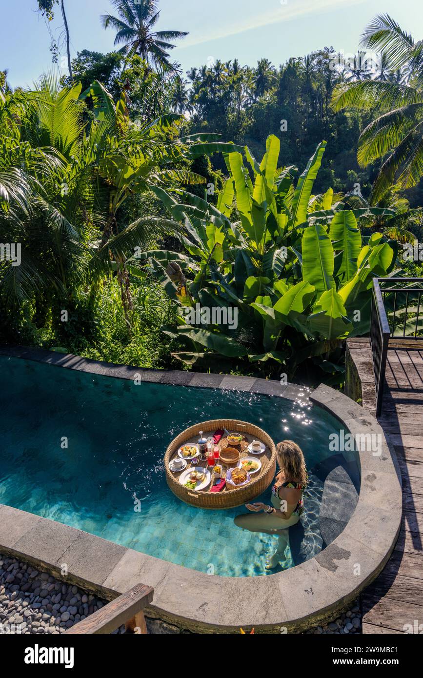 A young woman having breakfast on a floating basket in a swimming pool ...