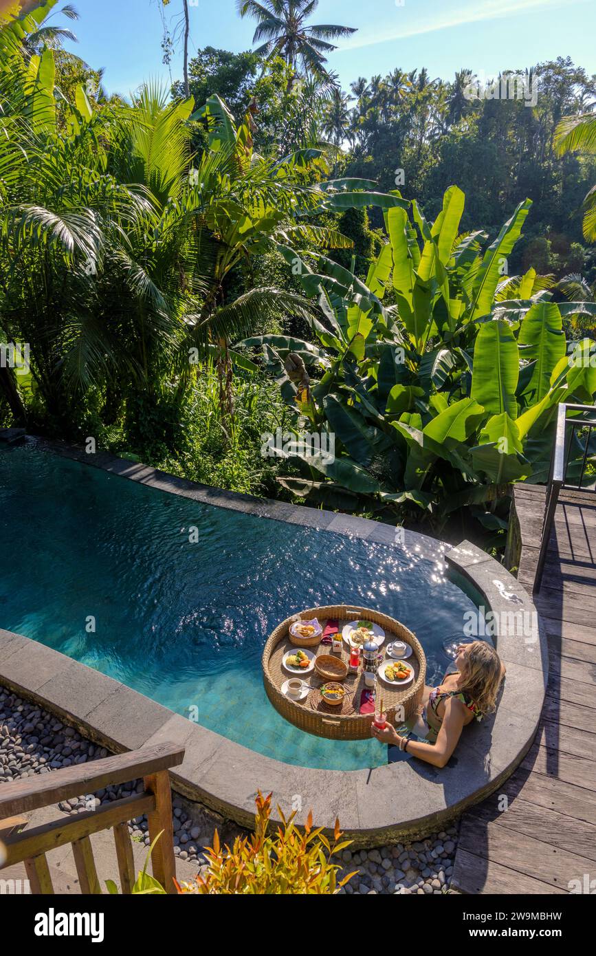 A young woman having breakfast on a floating basket in a swimming pool ...