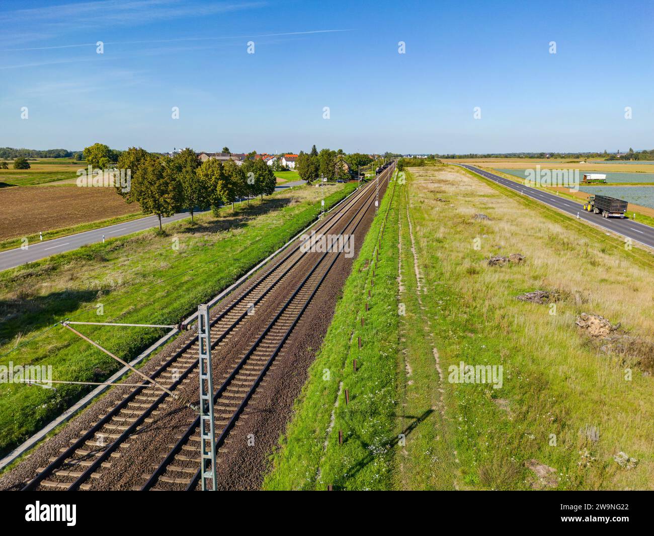 An electric railroad line with tracks next to two roads in a rural area ...