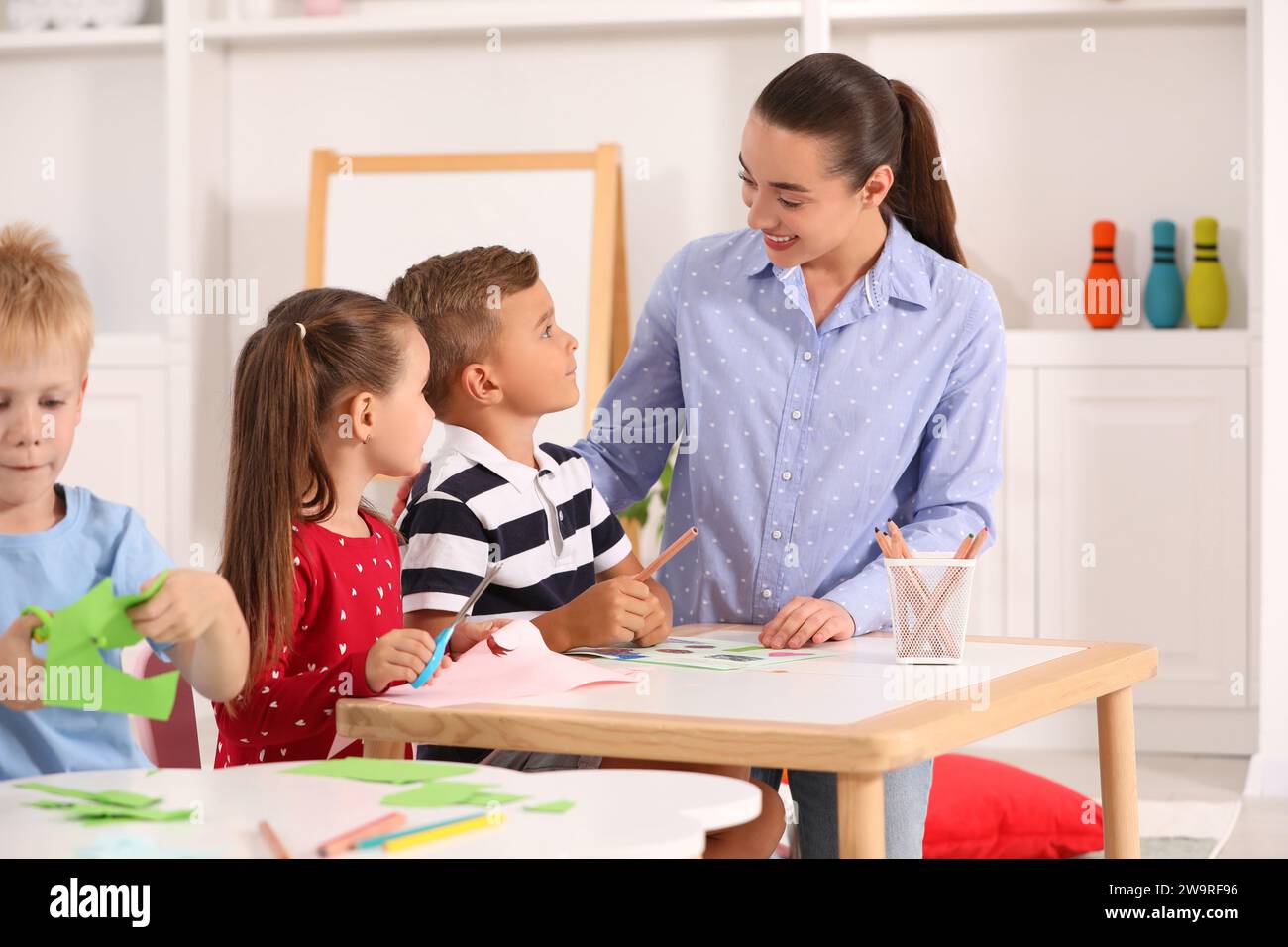 Nursery teacher and cute little children playing at desks in ...