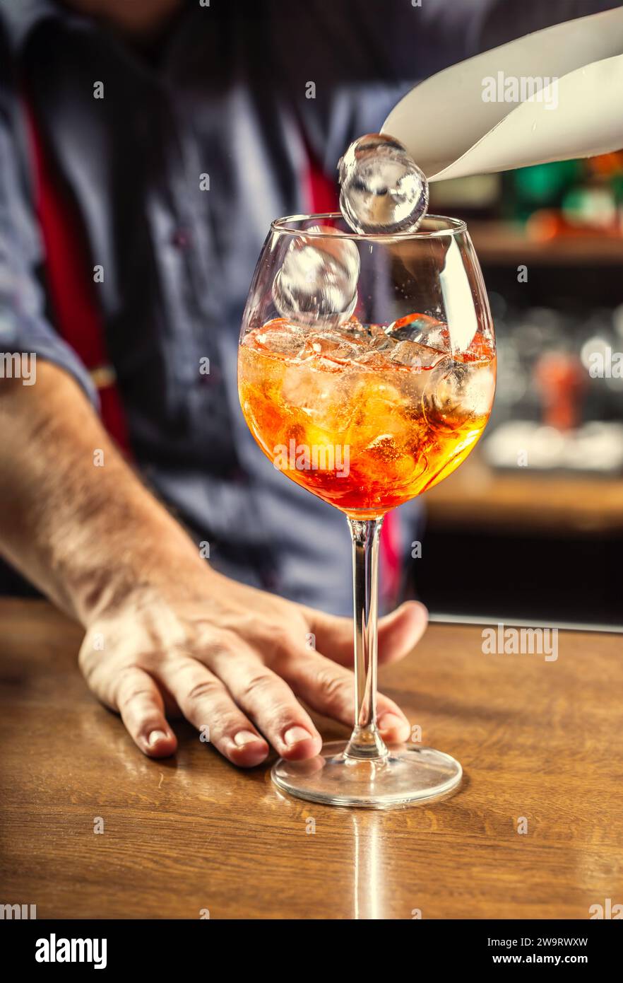 The bartender at the bar prepares the summer cocktail Spritz Veneziano, adds ice cubes to the ...