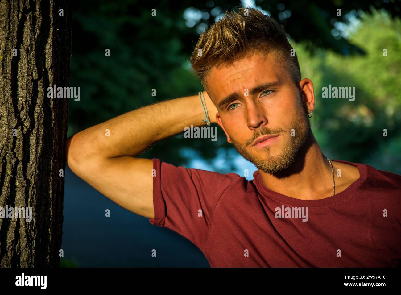 Sad, worried blond young man leaning against tree, outside in nature ...