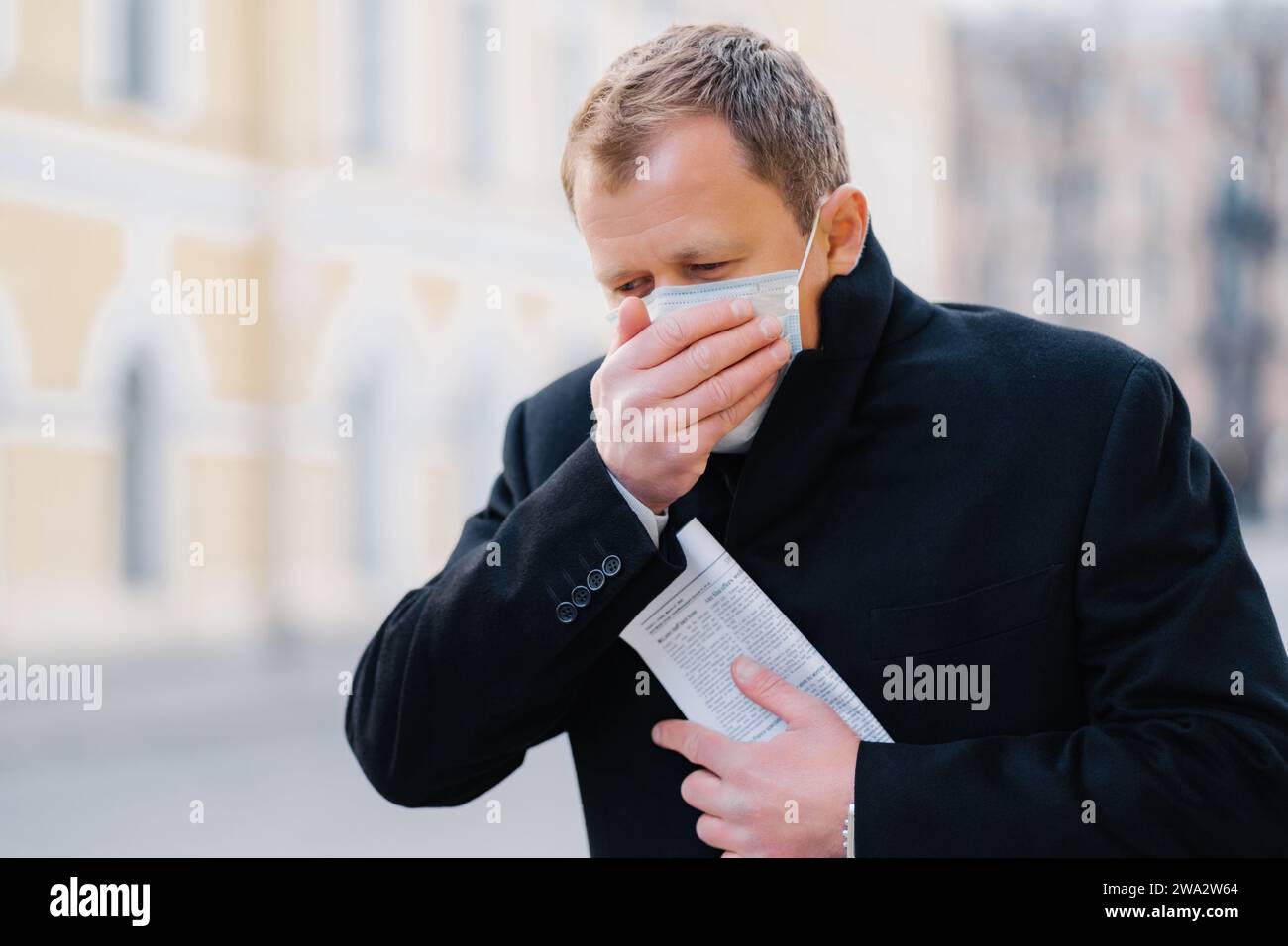 Sick man in black coat covering mouth with mask, outdoors, showing ...