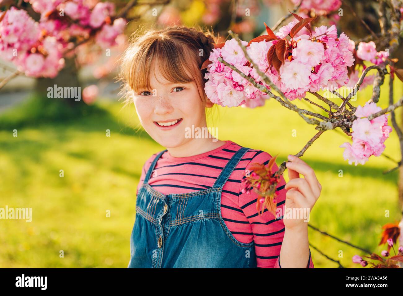 Outdoor spring portrait of a pretty little girl, standing between flowers of a japanese cherry ...