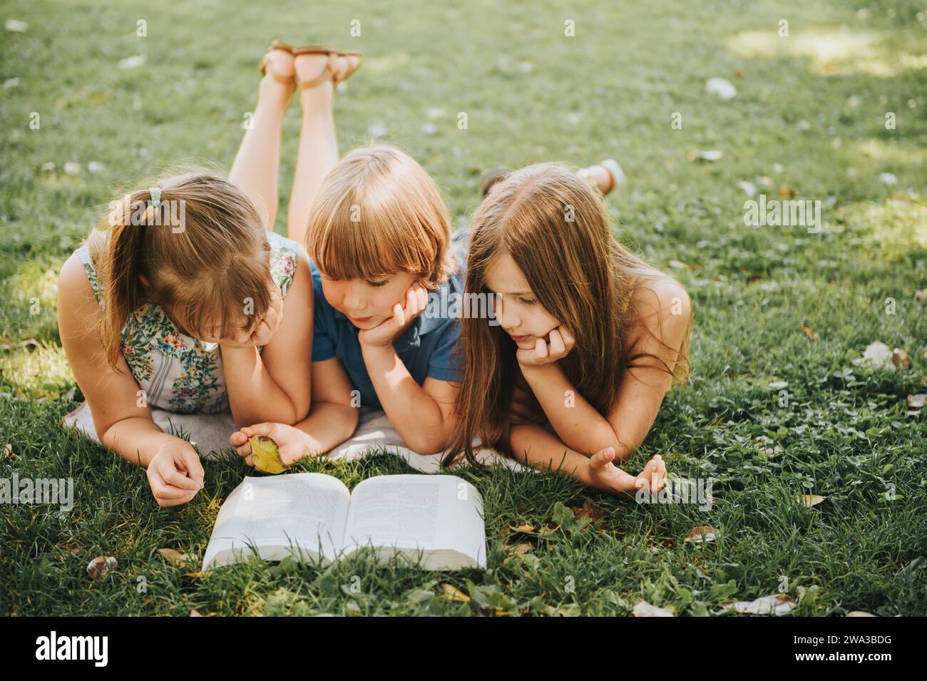 Group of three kids lying on green grass and reading story book ...