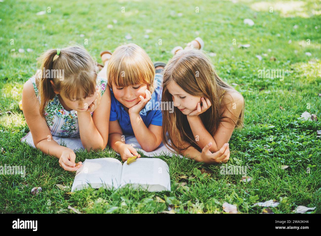 Group of three kids lying on green grass and reading story book ...