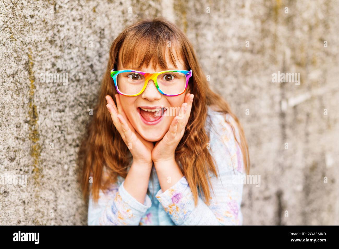 Little redhead girl wearing rainbow eyeglasses. Hands on cheeks looking ...